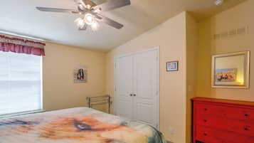 Um quarto com cama, ventilador de teto, janela com persianas, porta, cômoda e decoração nas paredes.