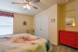 Um quarto com cama, ventilador de teto, janela com persianas, porta, cômoda e decoração nas paredes.
