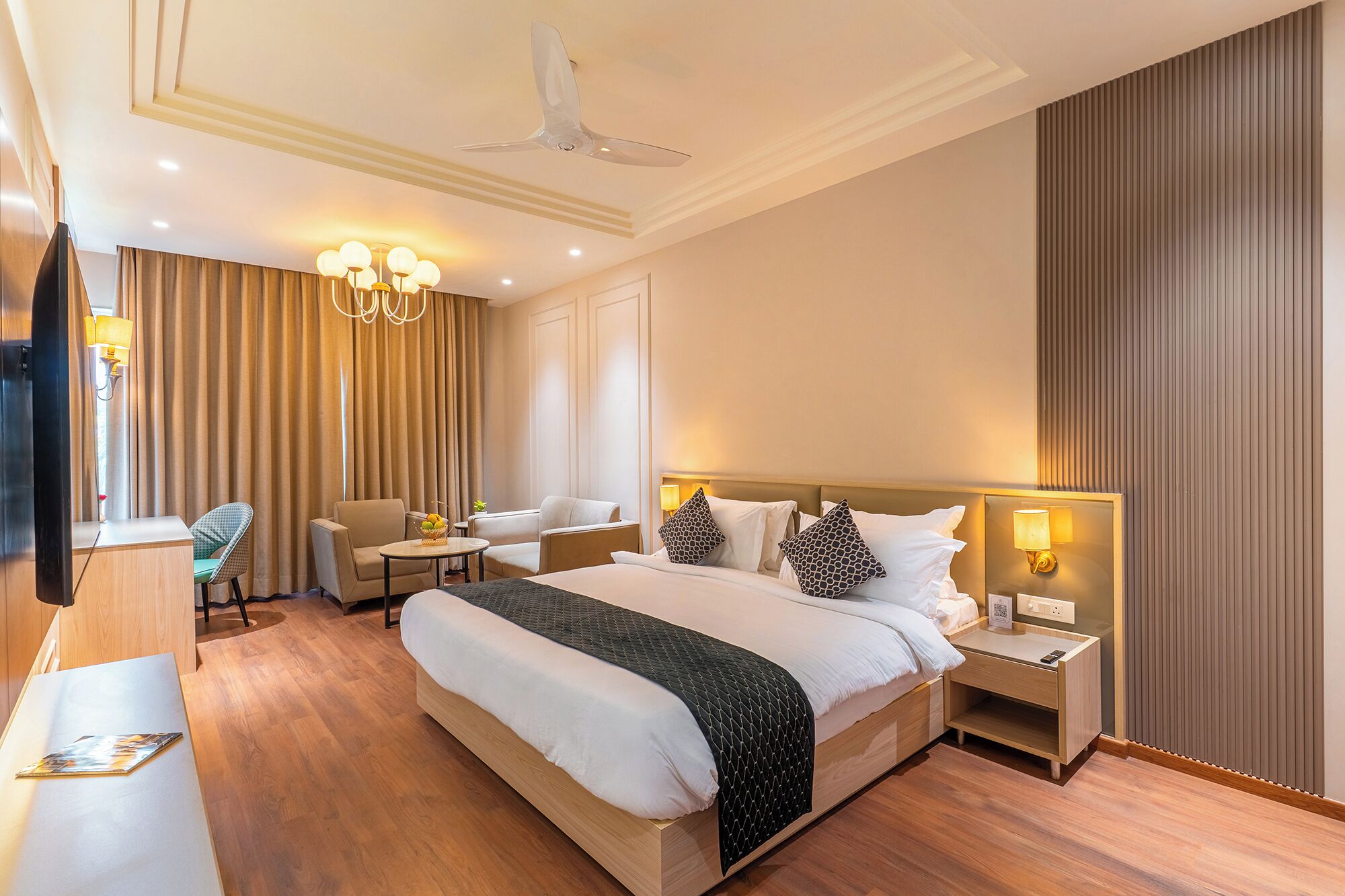 Quarto de hotel com uma cama grande, mesas de cabeceira, uma escrivaninha, uma cadeira, uma televisão e um ventilador de teto.