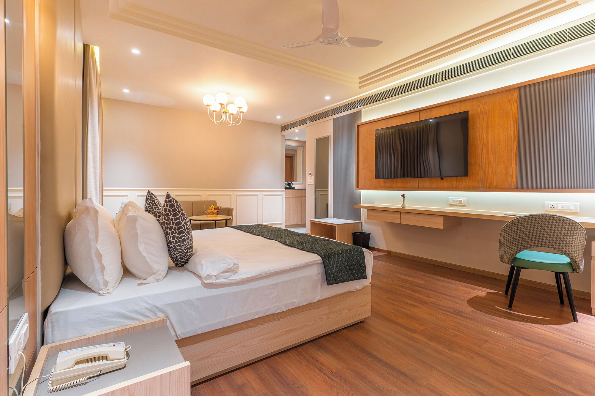 Quarto de hotel com cama, mesa de cabeceira, cadeira, escrivaninha, espelho e televisão.