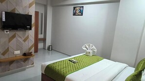 Quarto de hotel com uma cama, televisão presa à parede, uma pequena estante e um quadro na parede.