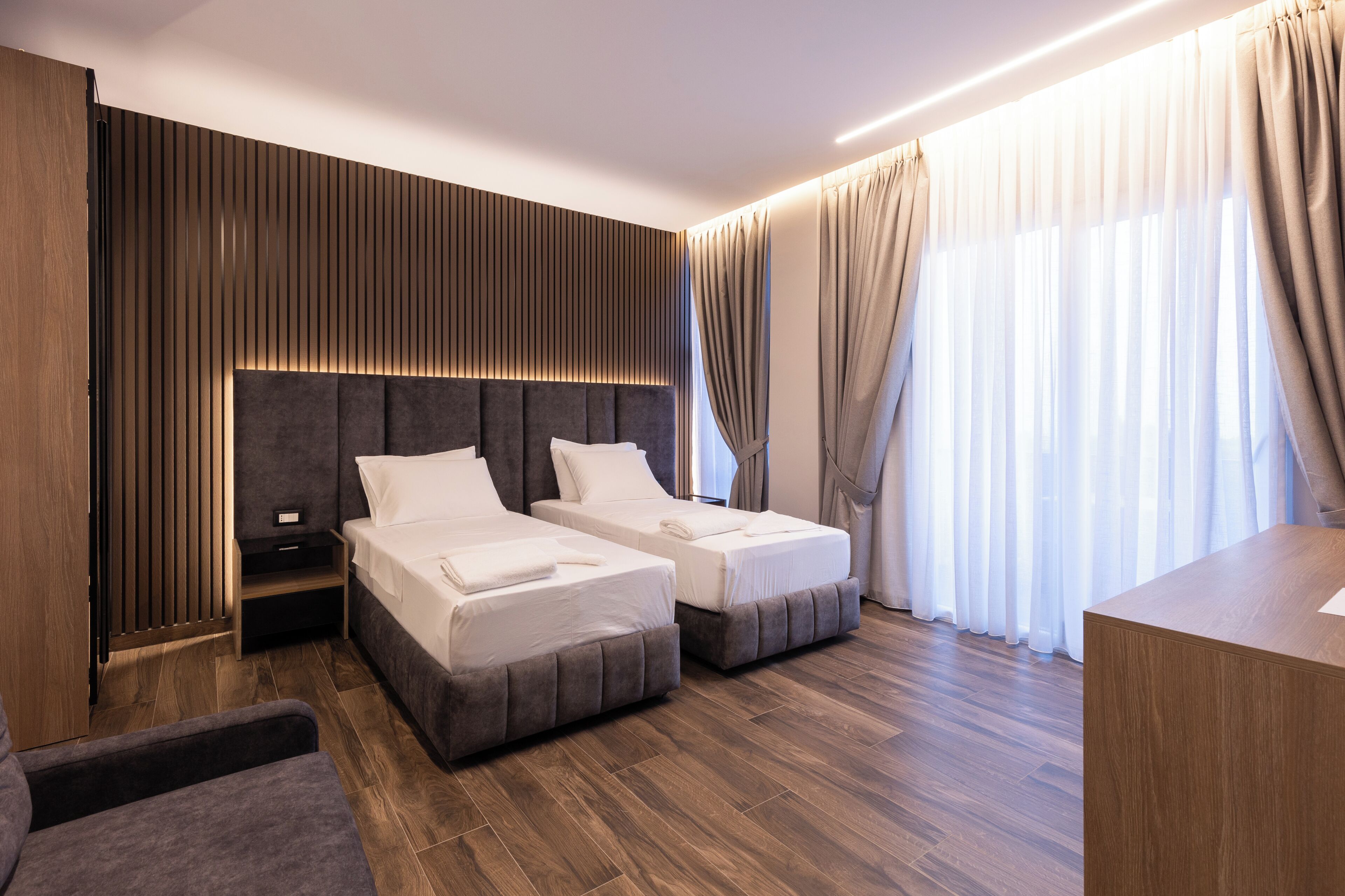 Quarto de hotel moderno com duas camas, painel de parede de madeira, escrivaninha de madeira e uma janela grande com cortinas.