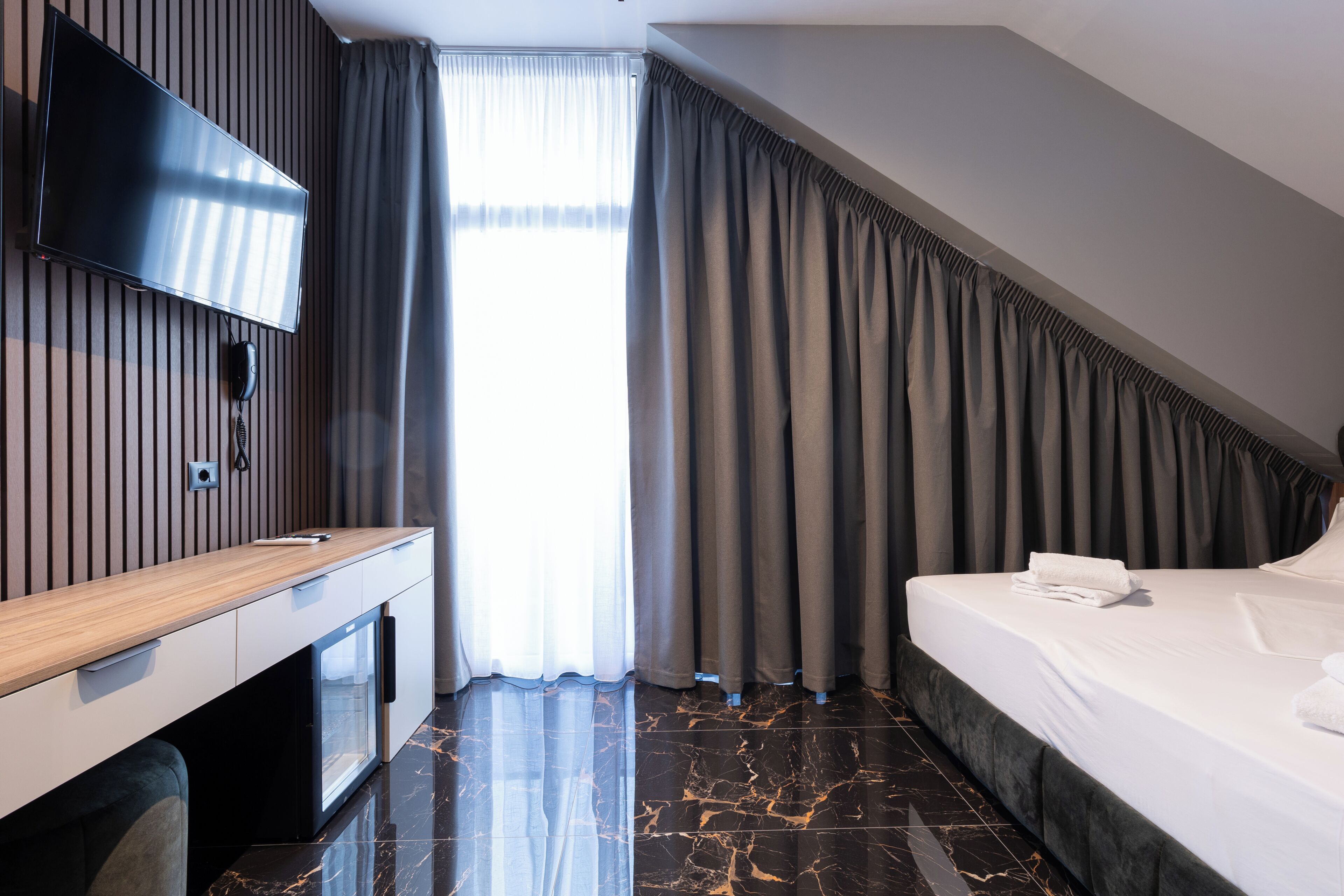 Quarto de hotel moderno com televisão de tela plana, escrivaninha de madeira, cama com roupa branca e uma grande janela com cortinas.