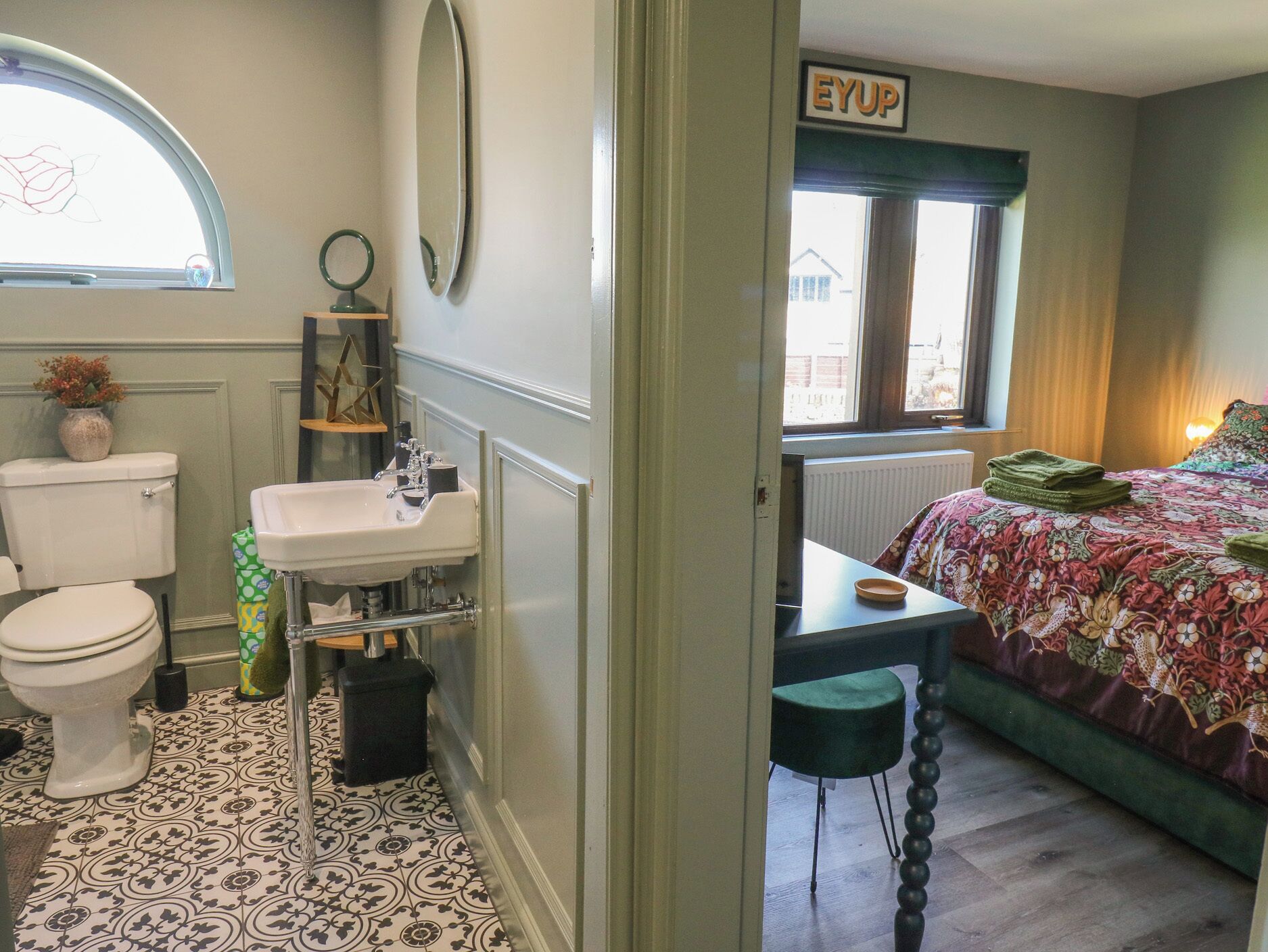 Casa de banho com sanita, lavatório e espelho, uma pequena janela com vista e uma cama com colcha estampada.
