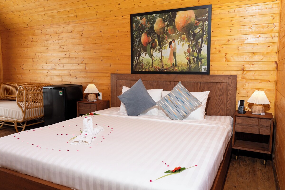 Uma cama bem arrumada com almofadas e um cobertor, uma cabeceira de madeira, uma pequena mesa de cabeceira com um abajur e uma cadeira de vime.