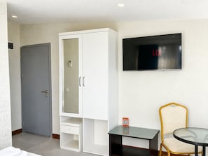 Quarto com porta cinza, guarda-roupa branco, uma pequena mesa com cadeira e televisão fixada na parede.