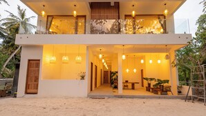 Uma casa moderna de dois andares com varanda, várias luzes penduradas e chão de areia.