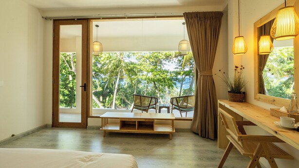 Quarto com cama, escrivaninha de madeira, duas cadeiras, janela com cortinas e vista para árvores.