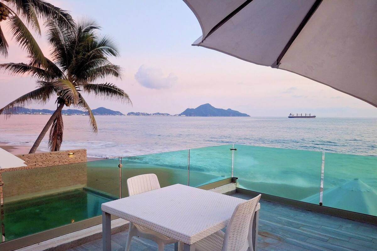 Uma piscina infinita com água cristalina, uma mesa e cadeira brancas, uma palmeira e vista para o mar e ilhas ao longe.