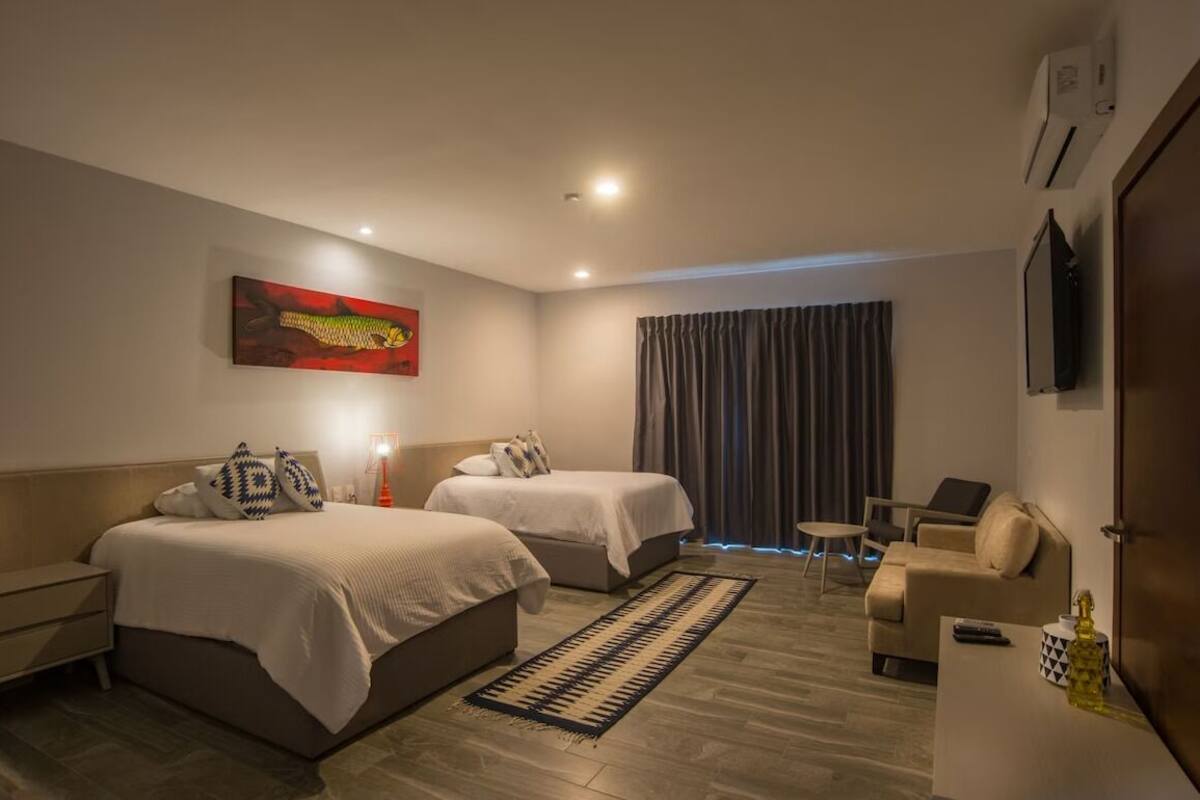 Quarto de hotel com duas camas, um sofá, uma televisão e uma janela com cortinas.