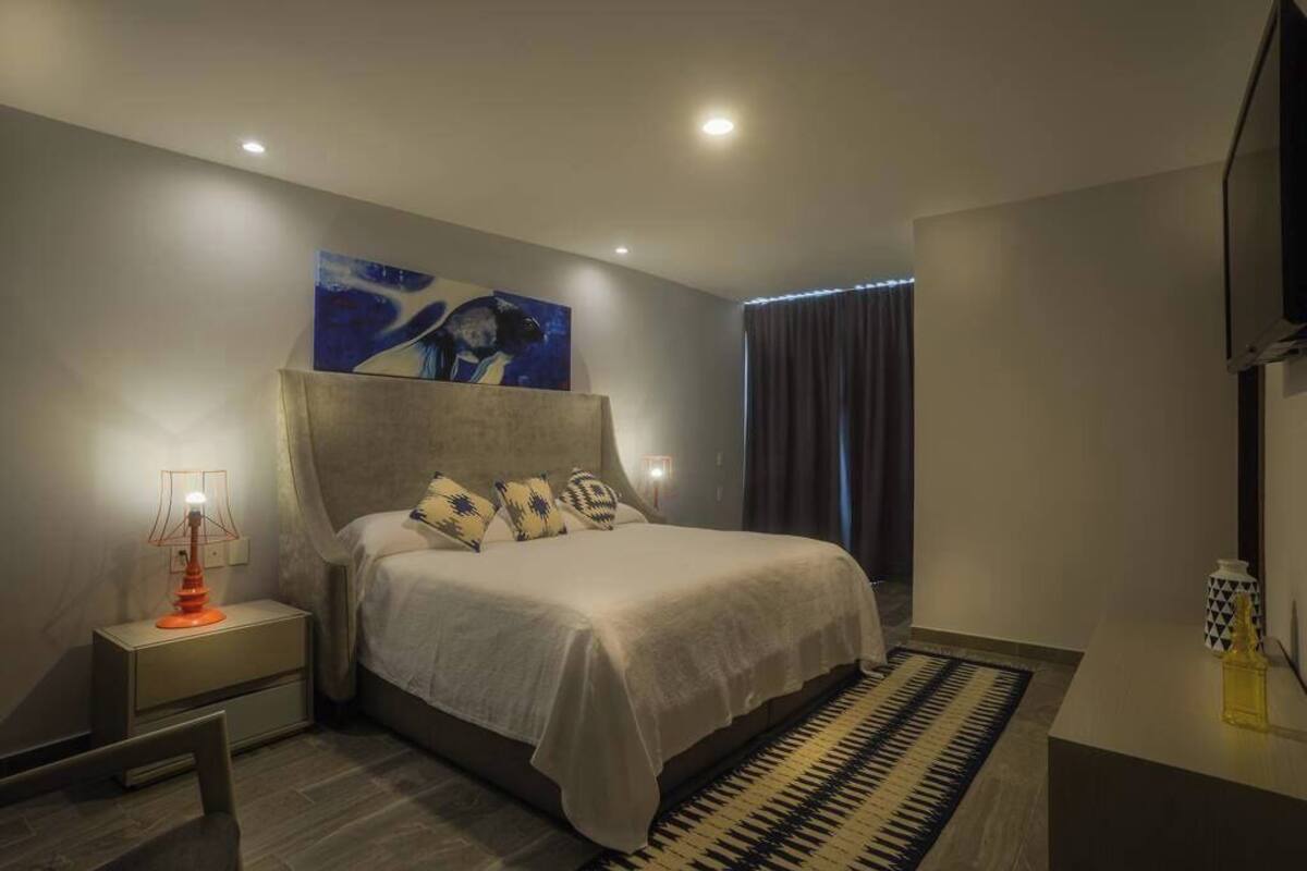 Quarto de hotel moderno com uma cama grande, uma mesa de cabeceira com abajur, um quadro na parede e vista para o exterior.
