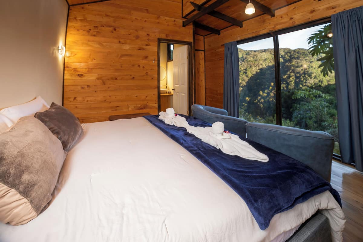 Quarto de cabana de madeira com cama, uma cadeira e uma janela grande com vista para a floresta.