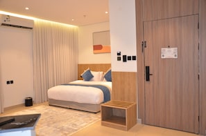 Quarto de hotel moderno com uma cama, uma porta de madeira, um banquinho de madeira pequeno, um ar condicionado fixado na parede e uma obra de arte emoldurada.