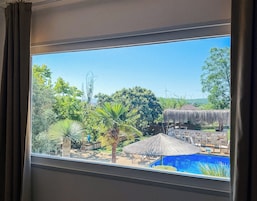 Vista da janela de uma área de piscina com estrutura de palha, palmeiras e céu claro.