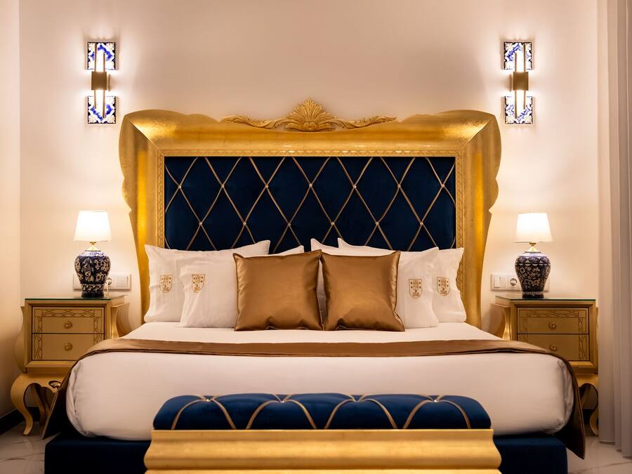 Uma cama luxuosa com cabeceira dourada, almofadas brancas e castanhas, e mesas de cabeceira combinando com abajures.
