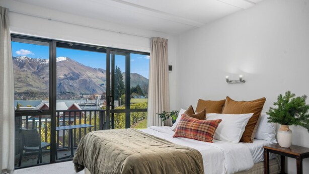 Quarto com cama grande, mesa de cabeceira, planta em vaso e vista para montanhas através de uma varanda.