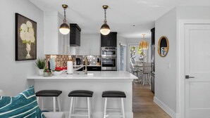 Cozinha moderna com armários brancos, uma ilha central com banquetas, eletrodomésticos de aço inoxidável e iluminação pendente.