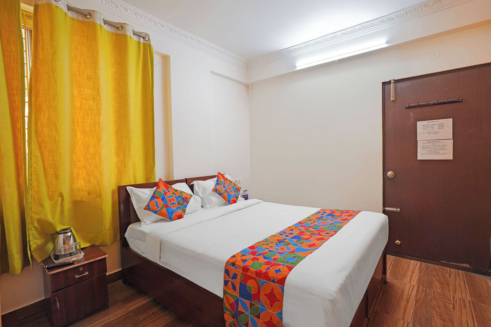 Quarto de hotel com cama, mesa de cabeceira, porta de madeira e cortina amarela.