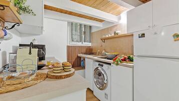 Uma cozinha compacta com máquina de lavar, teto de madeira e janela com persianas.
