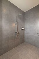 Área de duche com chuveiro manual, torneira fixada na parede e um pequeno espaço para produtos de higiene.