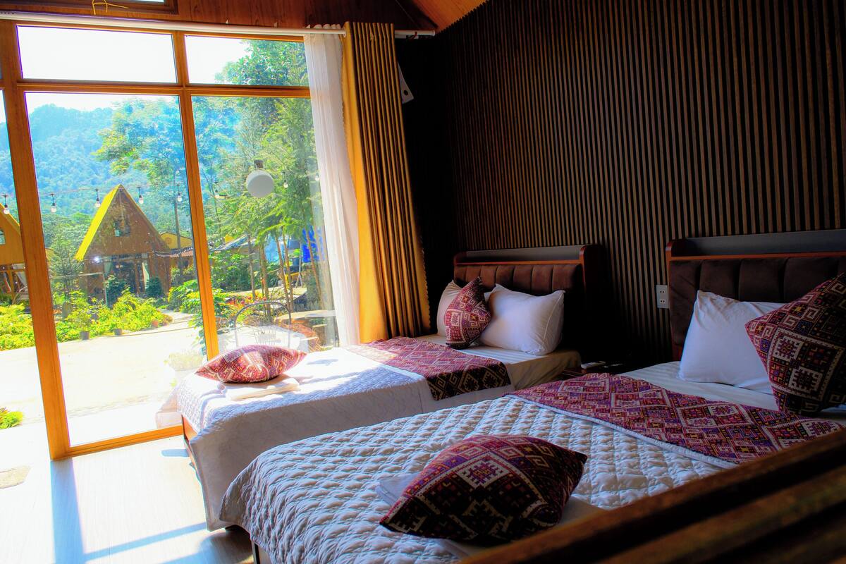 Quarto com duas camas, uma janela grande e vista para o verde e um prédio lá fora.