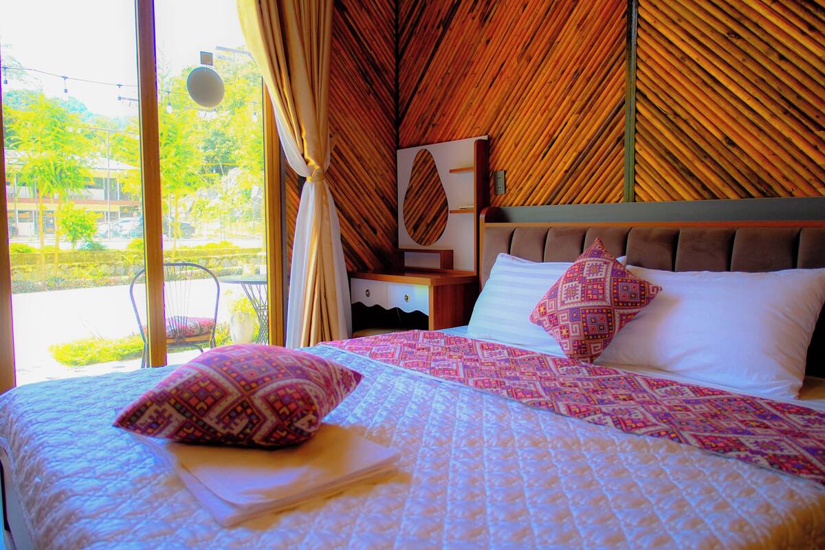 Cama com roupa de cama e almofadas estampadas, teto de madeira e vista para o verde lá fora.