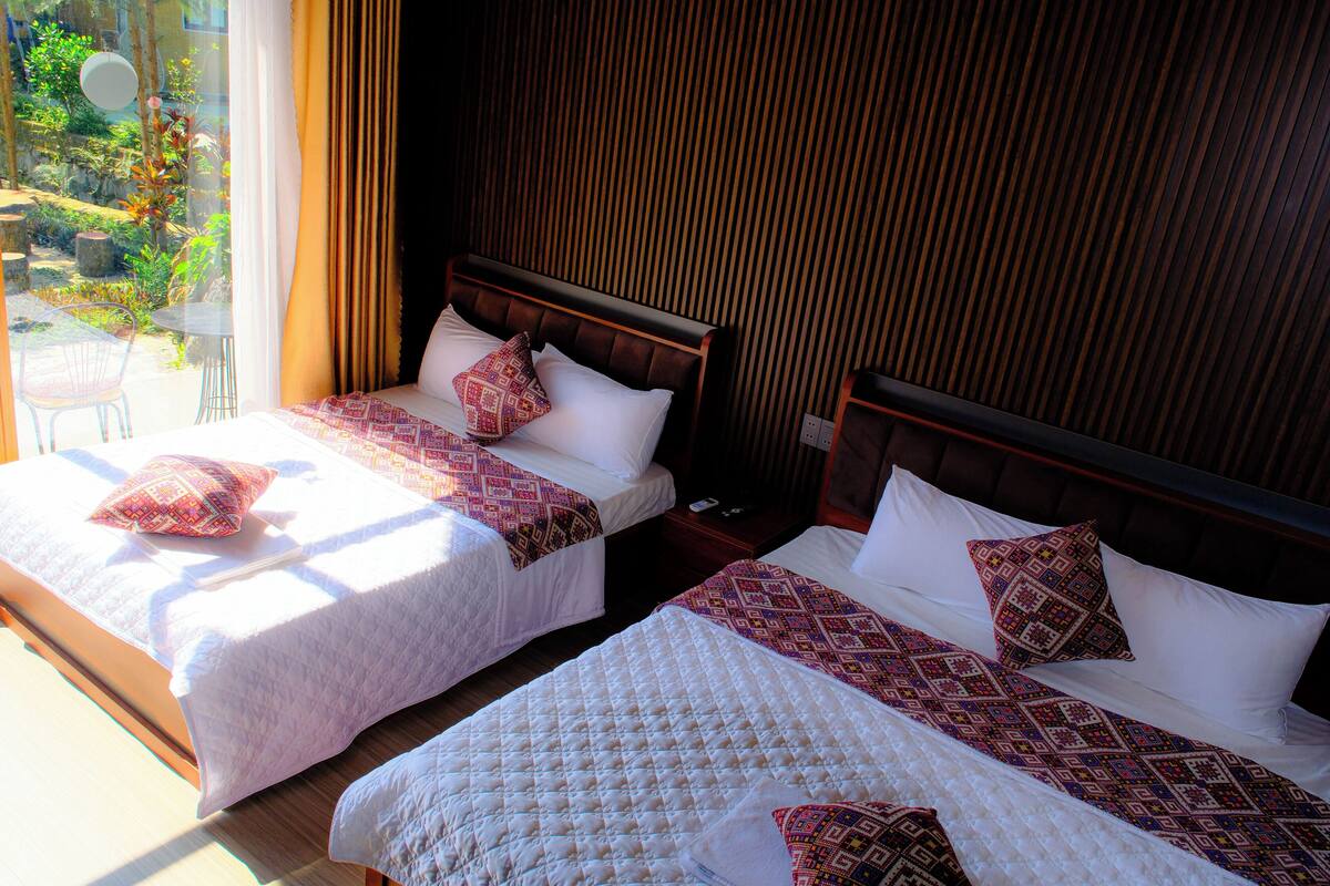 Duas camas com roupa de cama estampada, uma cabeceira de madeira e vista para área externa com plantas e uma mesa.