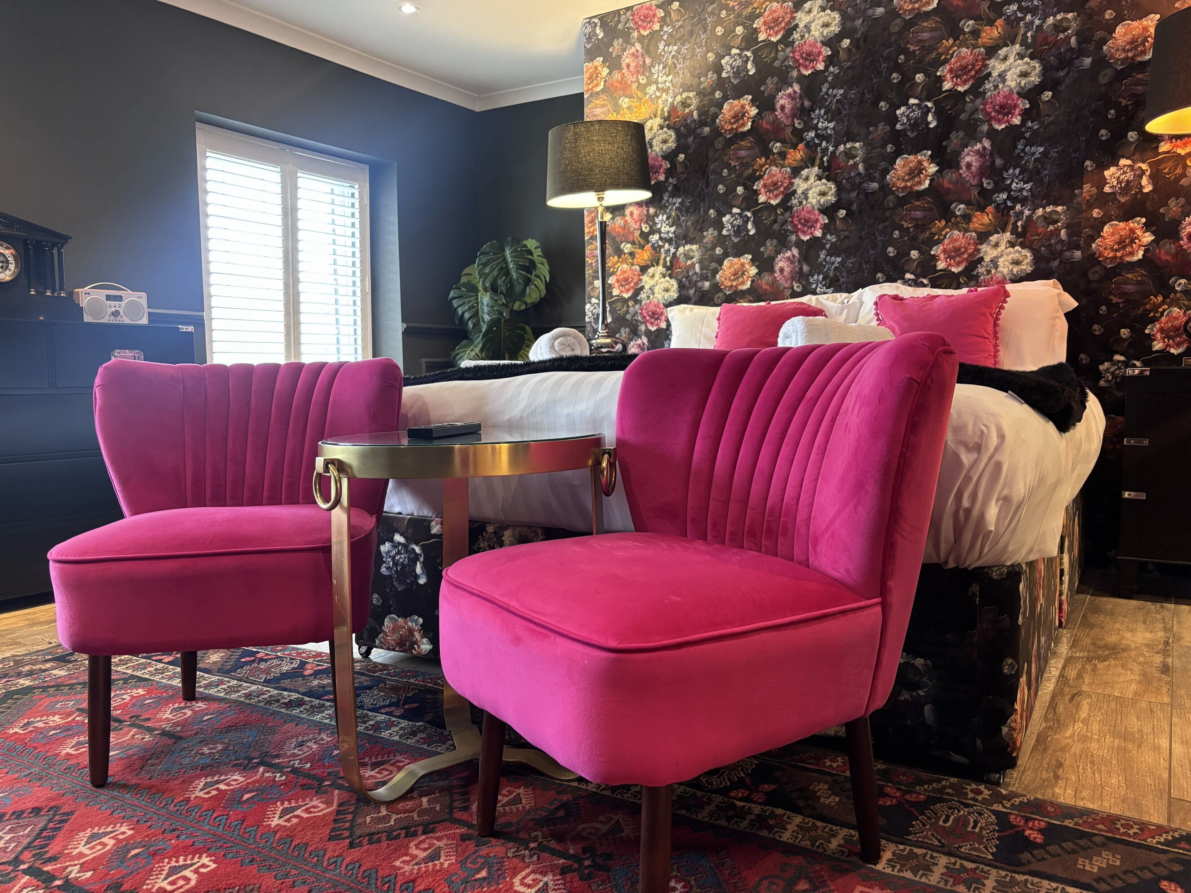 Um quarto com uma cama, duas cadeiras cor-de-rosa, uma mesinha redonda, um abajur e papel de parede floral.