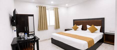 Quarto de hotel com cama, escrivaninha, cadeira, televisão e janela com cortinas.