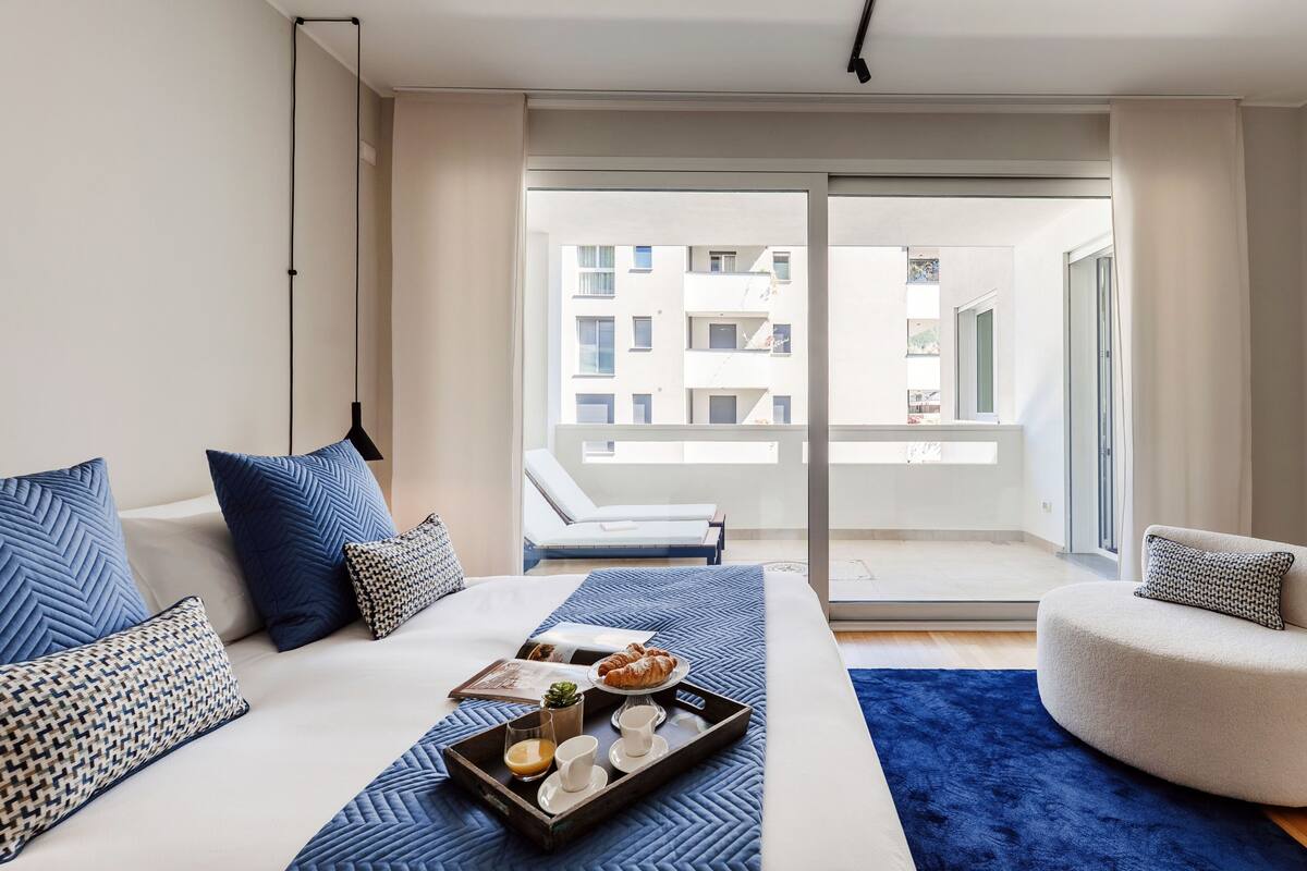 Quarto de hotel moderno com cama, bandeja de café da manhã, sofá e varanda com vista para a cidade.