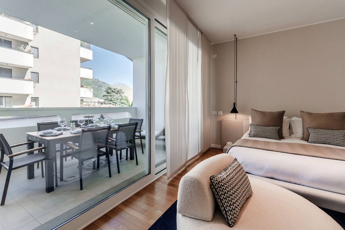 Quarto de hotel moderno com varanda, área de refeições com cadeiras e mesa, e cama com almofadas.