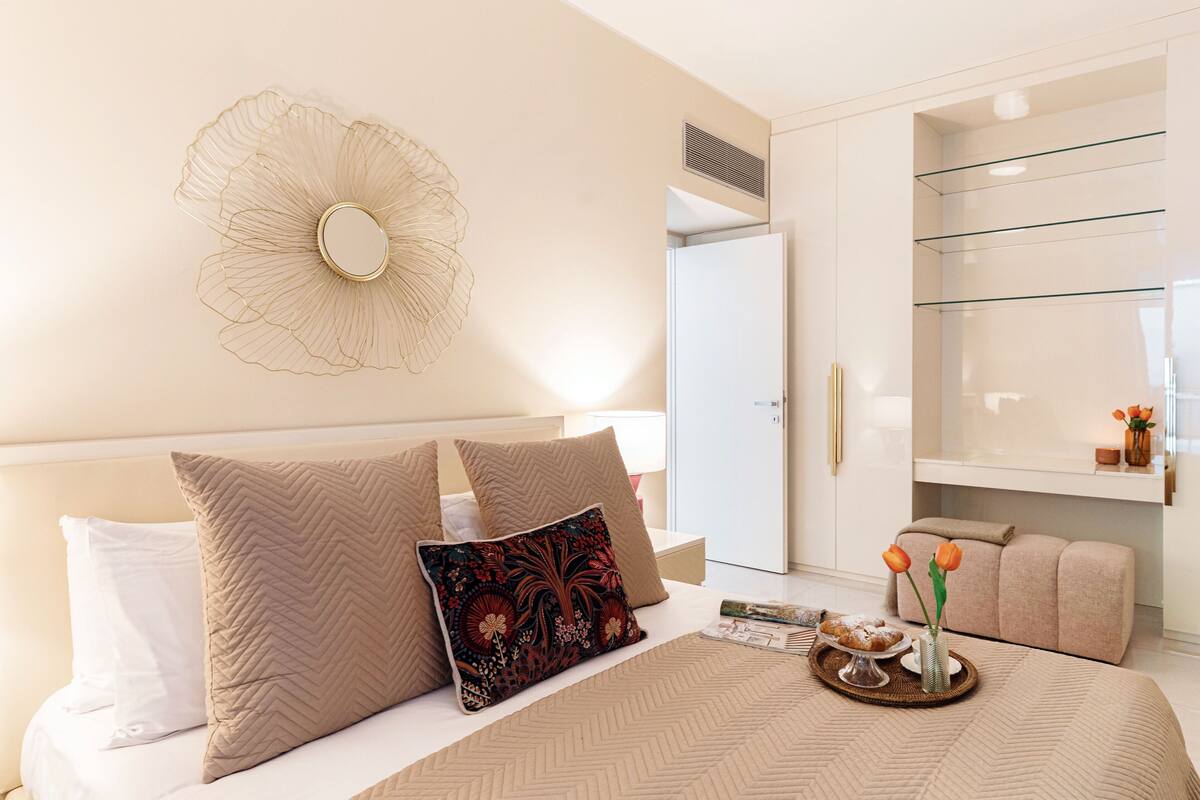 Quarto de hotel com cama, um painel decorativo, um aparador de vidro e uma bandeja com flores e doces.