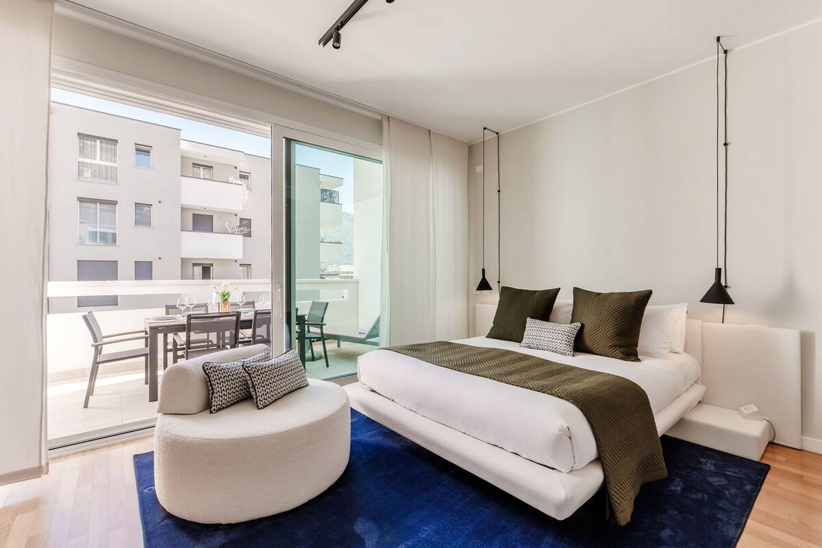 Quarto de hotel moderno com cama, área de estar circular, varanda com mesa de jantar e vista para a cidade.