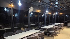 Restaurante com mesas e cadeiras dispostas no interior, grandes janelas e luminárias pendentes.