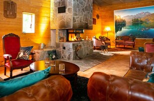 Interior acolhedor de um chalé, com lareira de pedra, móveis de couro e um quadro grande de um lago.