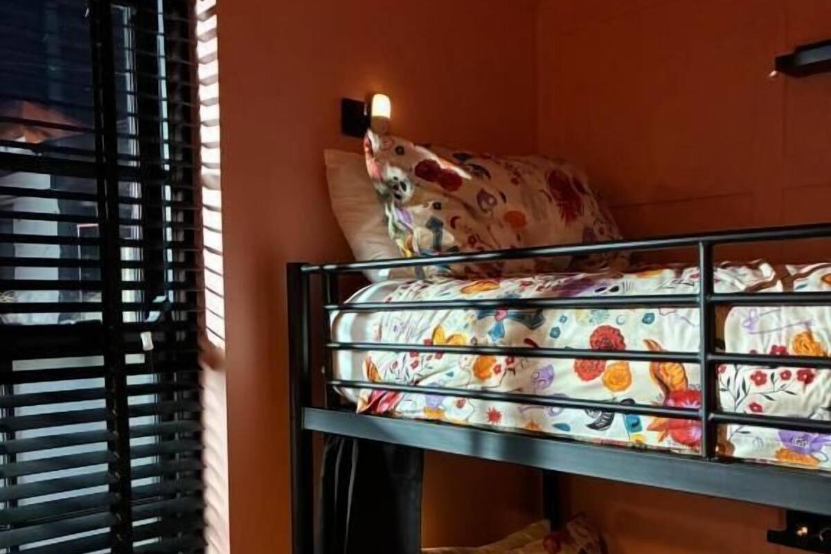 Uma cama beliche com roupa de cama florida, uma janela com persianas e uma prateleira fixada na parede.