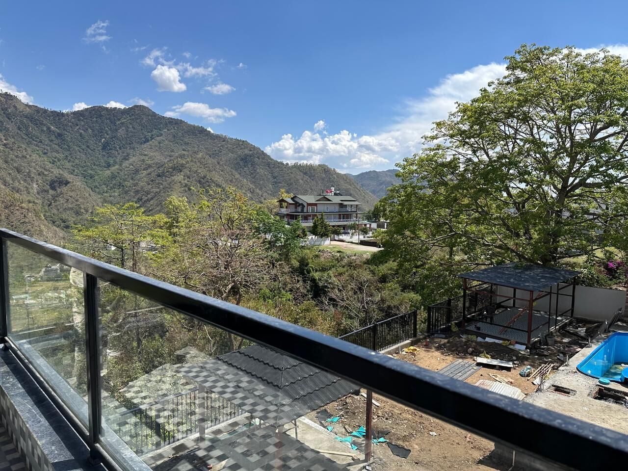 Vista de montanha de uma varanda com guarda-corpo de vidro, um edifício pequeno e uma árvore.
