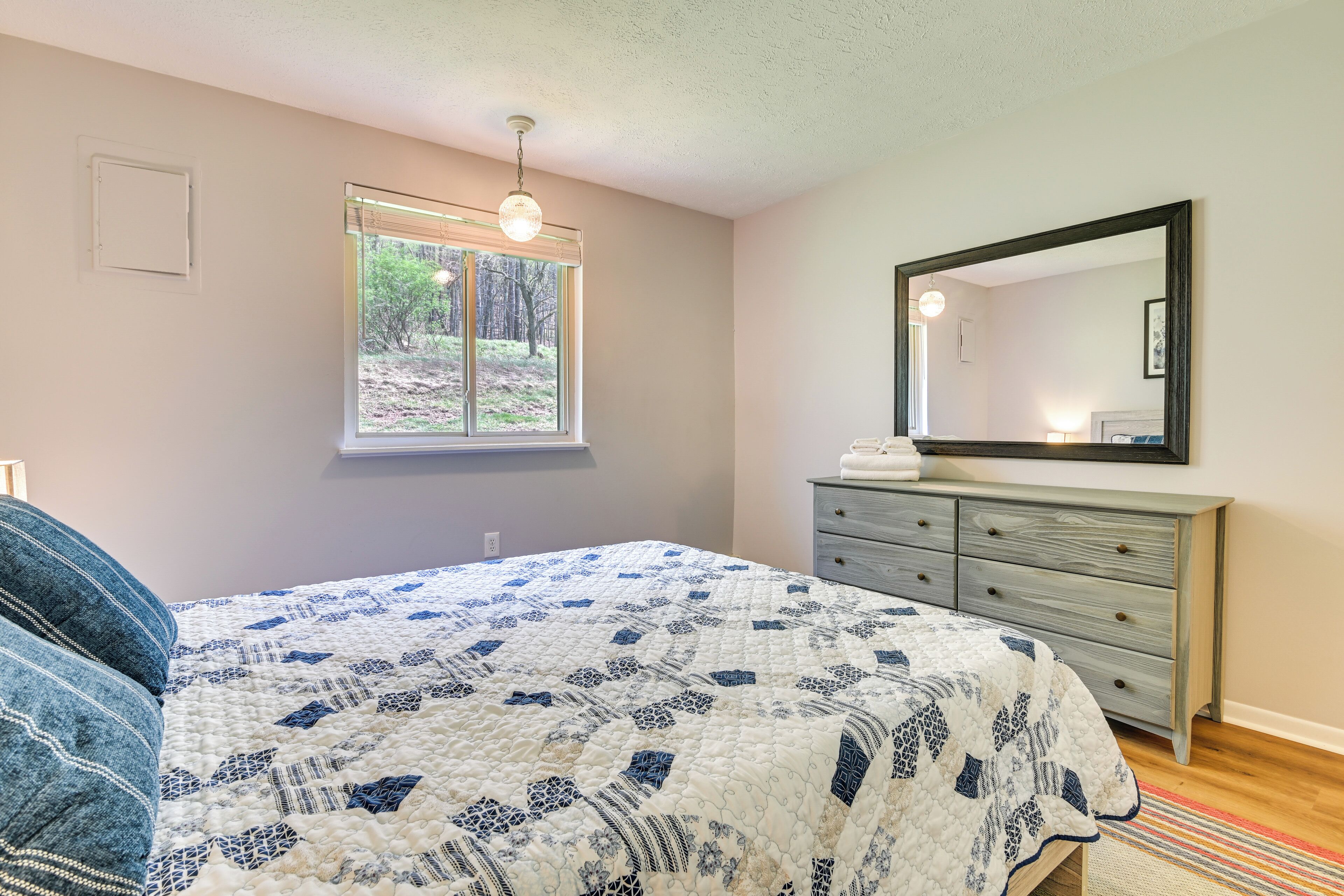 Quarto com cama, cômoda, espelho e janela com vista para árvores.