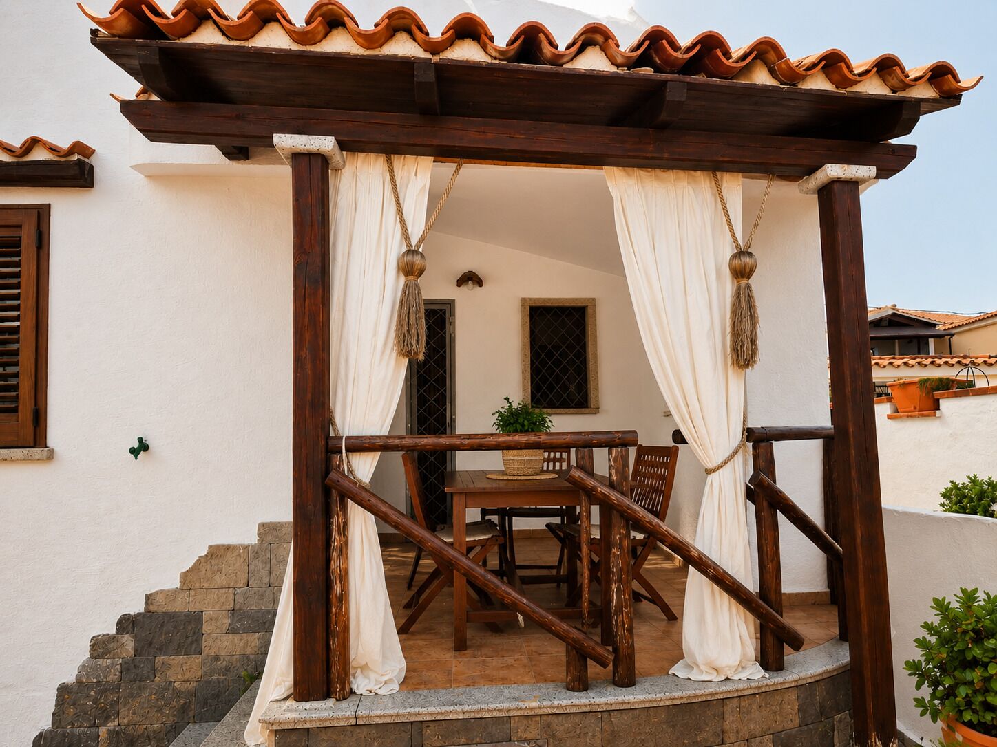 Uma pérgola de madeira com cortinas brancas, uma mesa e cadeiras em frente a um edifício.