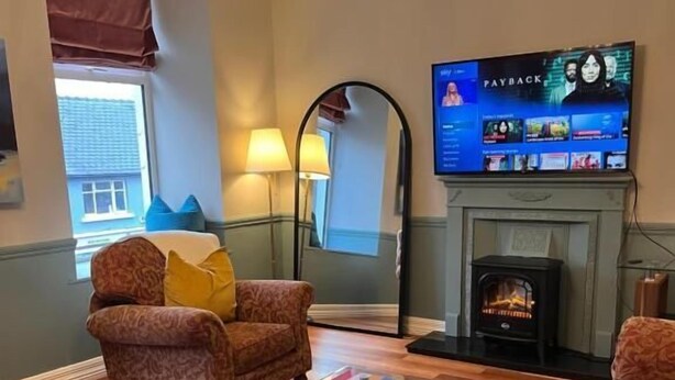 Sala de estar com sofás estampados, mesa de centro de vidro, lareira e televisão exibindo um filme.