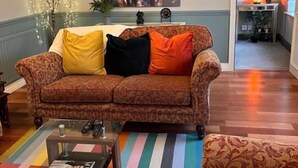 Uma sala de estar com um sofá estampado, uma mesa de centro com revistas, um tapete colorido e uma grande pintura abstrata na parede.