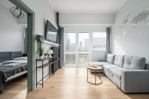 Uma sala de estar moderna com um sofá cinzento, uma mesa redonda pequena, uma cama com roupa de cama cinza, uma planta numa prateleira e uma janela grande com vista para edifícios.