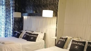 Quarto de hotel com duas camas, cada uma com roupa de cama branca e almofadas pretas com o logo Chanel.