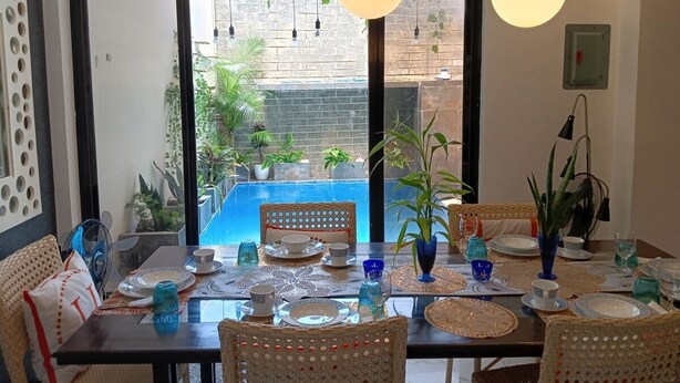 Uma sala de jantar com mesa posta para quatro pessoas, cercada por cadeiras de vime, com vista para uma parede de tijolos e plantas lá fora.