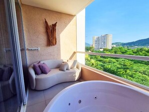 Uma varanda com sofá e banheira, um enfeite na parede, e vista para áreas verdes e edifícios.