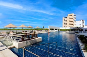 Piscina com espreguiçadeiras e guarda-sóis, um edifício alto ao fundo e céu azul claro.