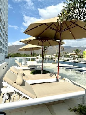Área de piscina com espreguiçadeiras e guarda-sóis, uma piscina e montanhas ao fundo.