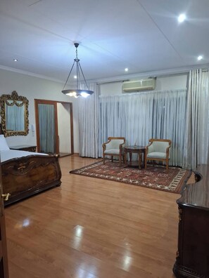 Quarto de hotel com cama de madeira, duas cadeiras, uma mesa pequena, ventoinha no teto e uma janela grande com cortinas.