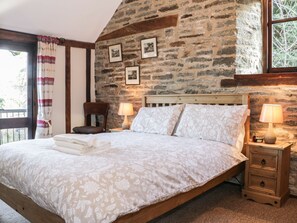 Quarto com parede de pedra, cama de madeira, duas mesas de cabeceira com luminárias, uma cadeira e uma janela com cortinas.