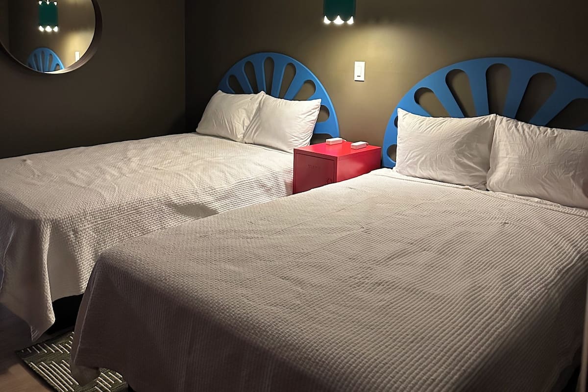 Quarto com duas camas, um espelho redondo, uma caixa vermelha e um lustre.